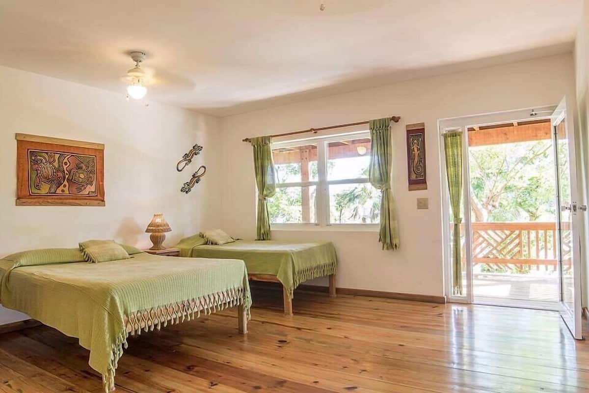 Quarto com duas camas, ventoinha no teto, janela com cortinas e porta que dá para o exterior.