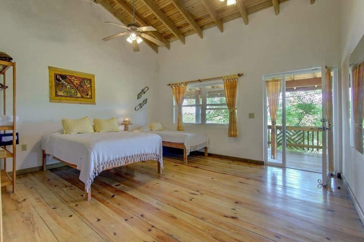 Quarto com duas camas, piso de madeira, ventilador de teto e janela com cortinas.