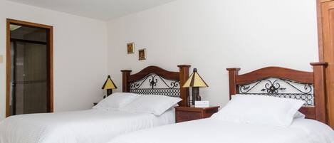 Quarto de hotel com duas camas de solteiro, cabeceiras de madeira, mesas de cabeceira com luminárias e porta para o banheiro.