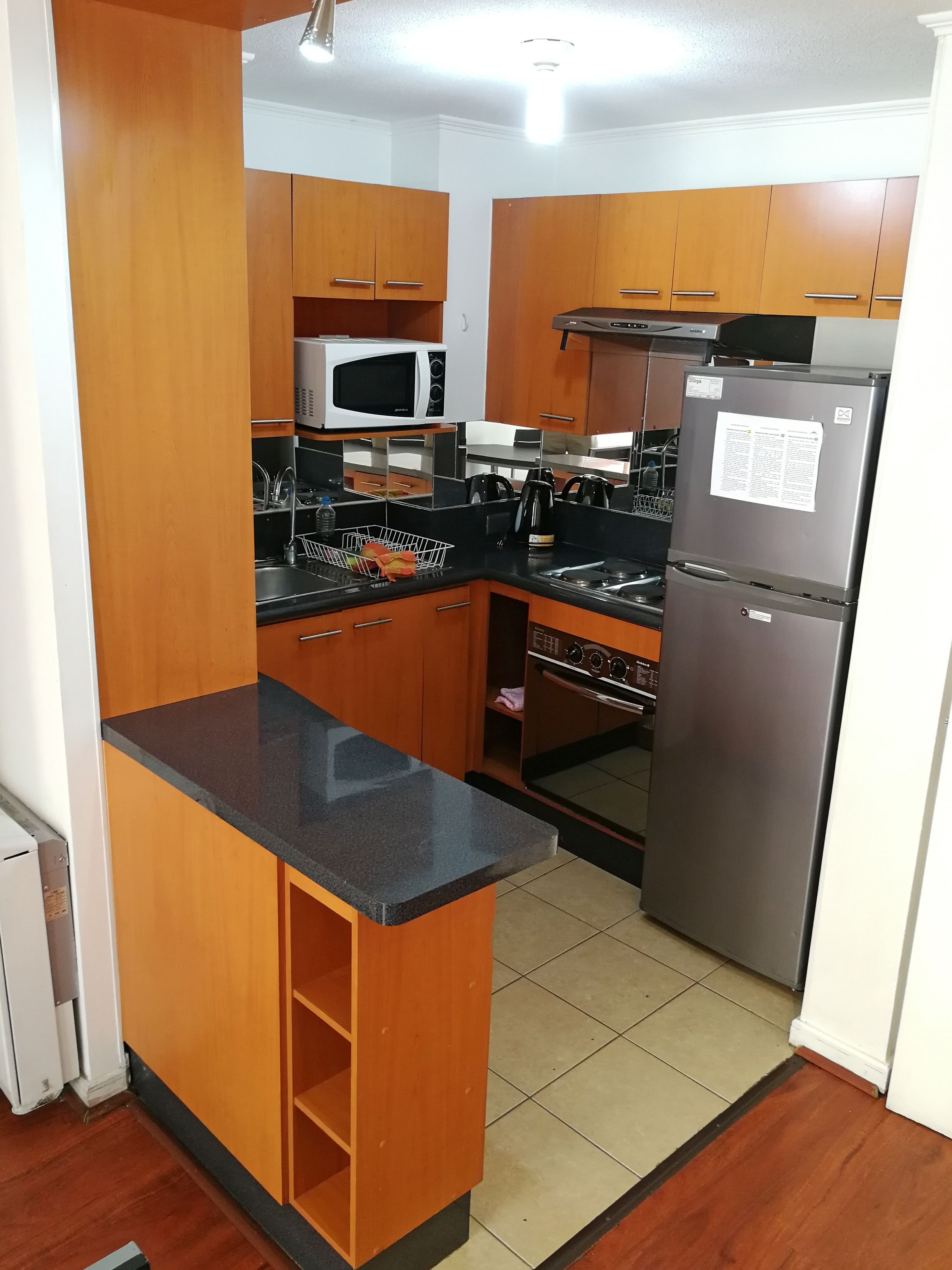 Cozinha compacta com armários de madeira, bancada preta, geladeira de aço inoxidável, micro-ondas e forno.