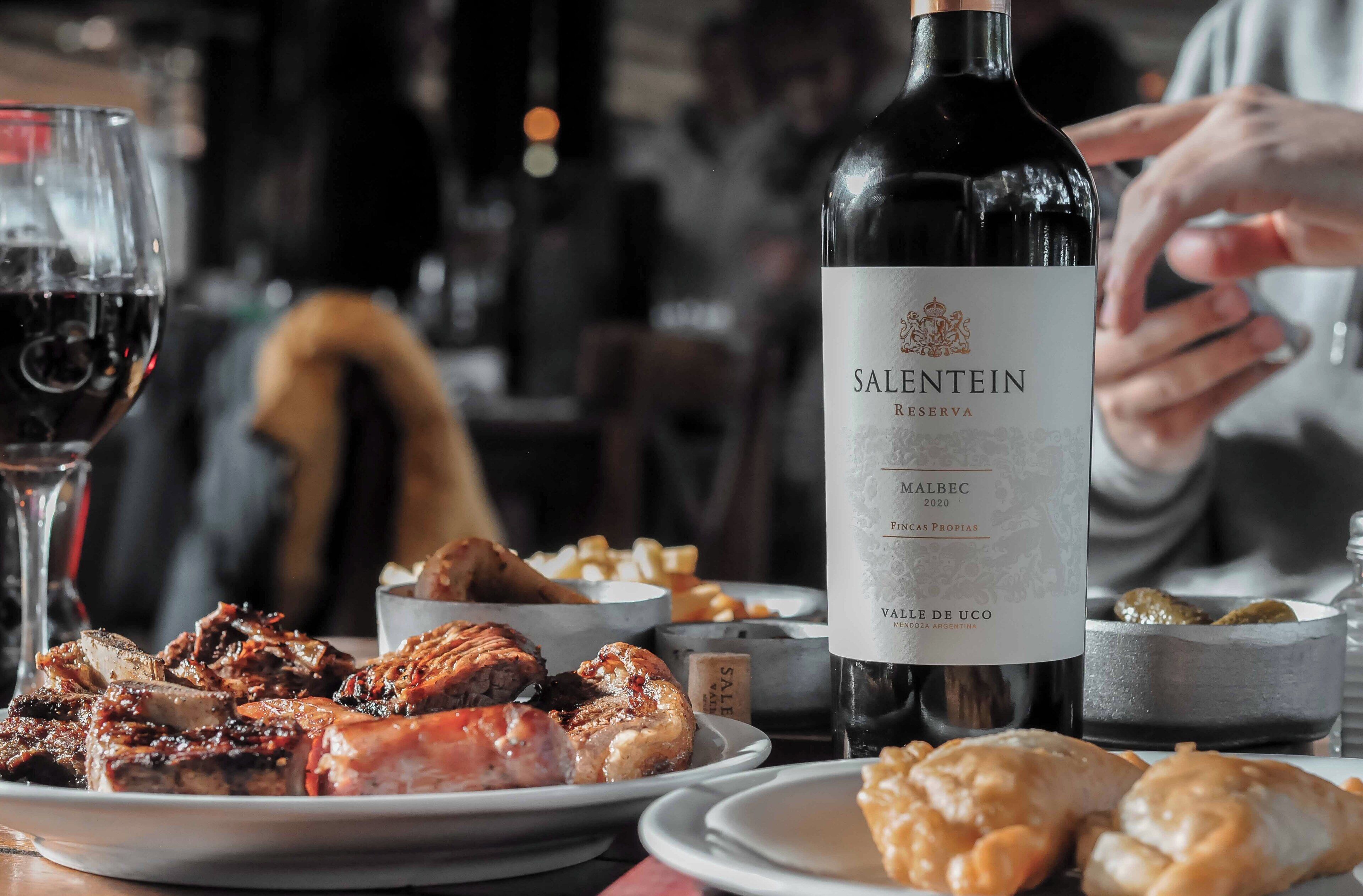 Uma garrafa de vinho tinto Salentein Reserva Malbec 2020, um prato com carne grelhada e uma taça de vinho tinto sobre uma mesa.