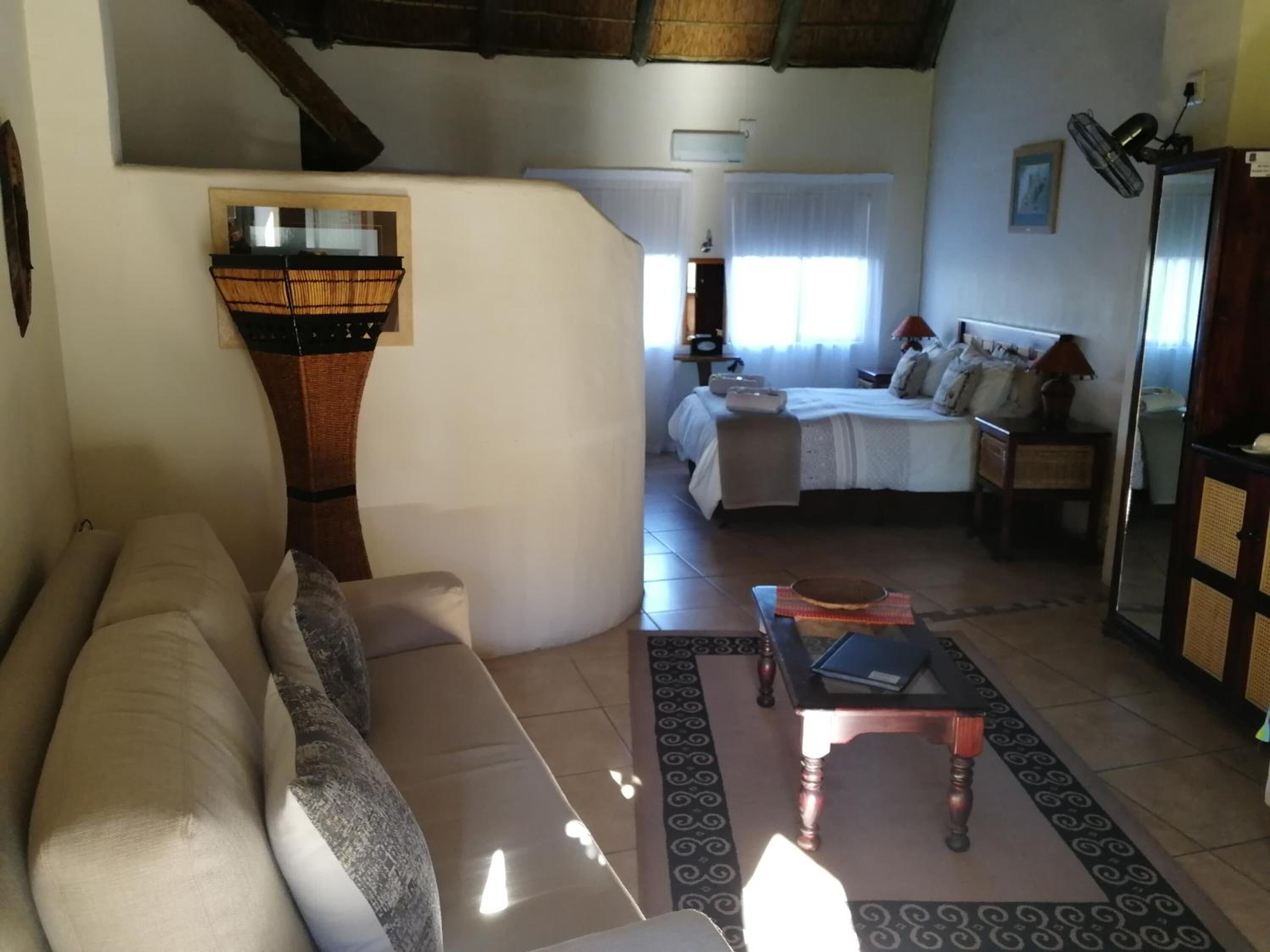 Uma sala de estar acolhedora, com um sofá, uma mesa de centro e um espaço de jantar. Há uma cama ao fundo, com uma mesa de cabeceira e um abajur.