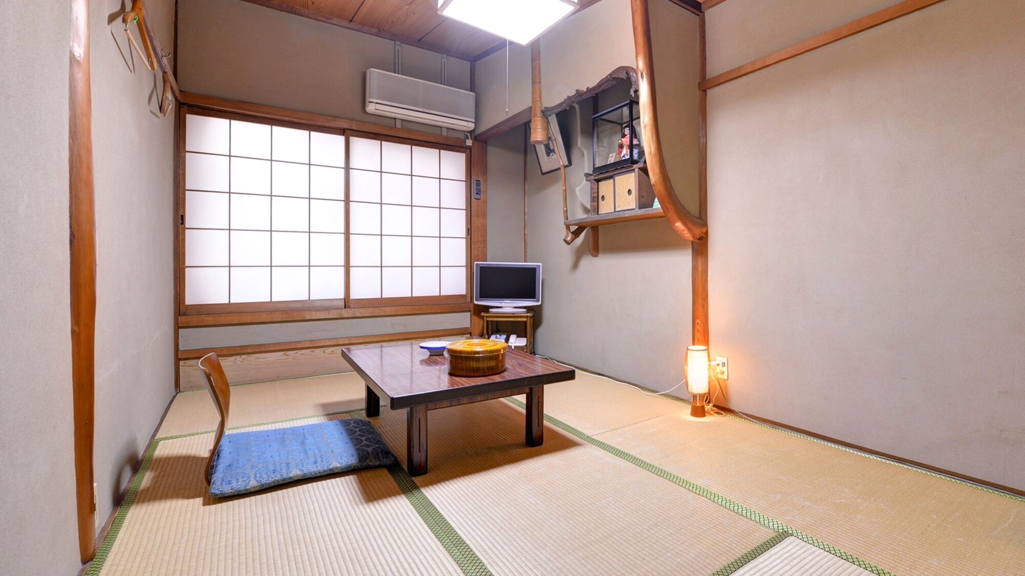 Um quarto japonês tradicional com piso de tatame, um tatame, uma mesa baixa com uma televisão e uma estante com objetos.