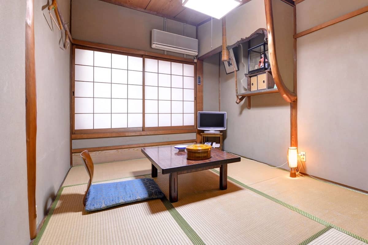 Um quarto japonês tradicional com piso de tatame, um tatame, uma mesa baixa com uma televisão e uma estante com objetos.