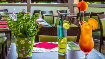 Mesa de restaurante com um balde de padrão floral verde, um copo alto com uma fatia de limão e canudo, e uma bebida colorida com um pedaço de laranja.