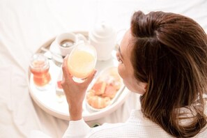 Uma pessoa sentada em uma cama com uma bandeja de itens de café da manhã, incluindo torradas, um copo de suco de laranja e uma chaleira.