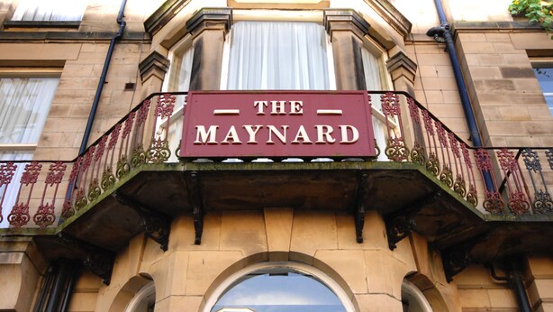 Um edifício com uma placa que diz "THE MAYNARD" acima de uma varanda com grades de metal ornamentadas.
