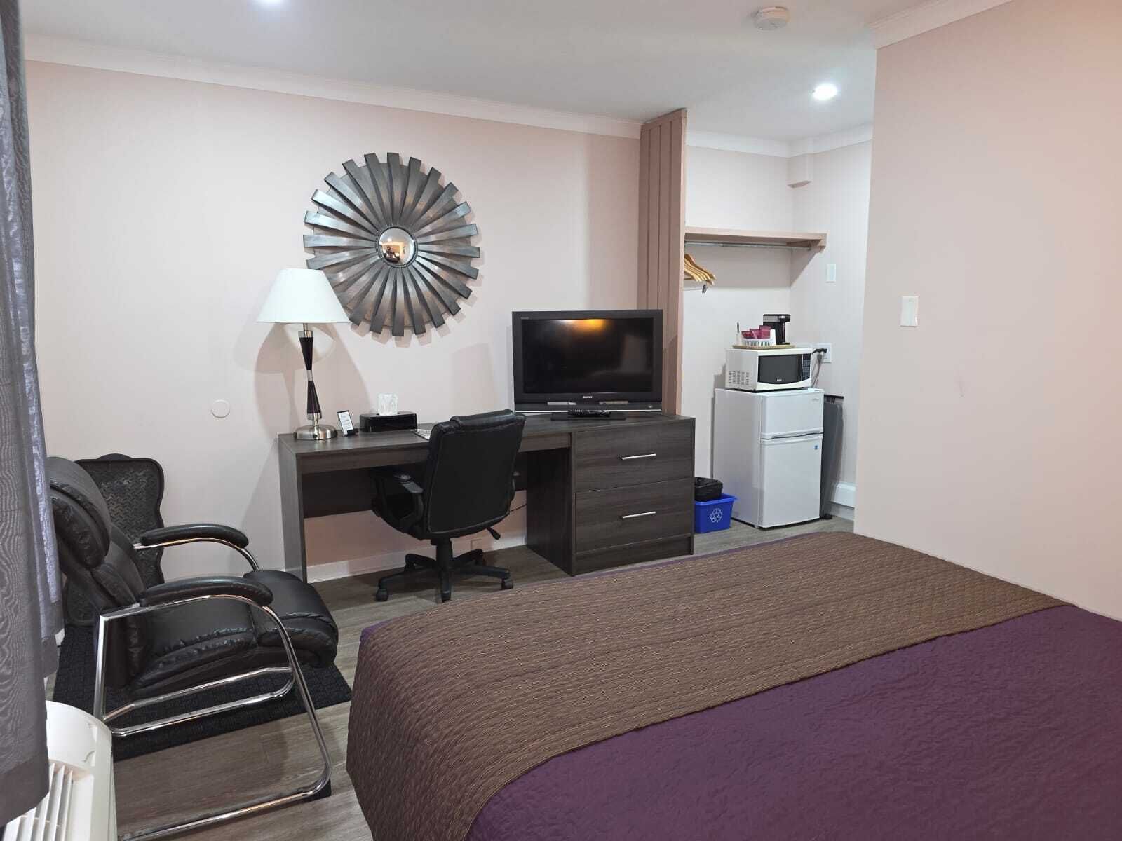 Quarto de hotel com cama, escrivaninha com computador, televisão, micro-ondas e um pequeno frigorífico.