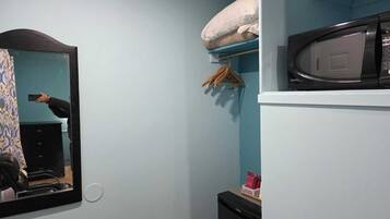 Quarto compacto com uma cama, cômoda, espelho, micro-ondas e um pequeno refrigerador.