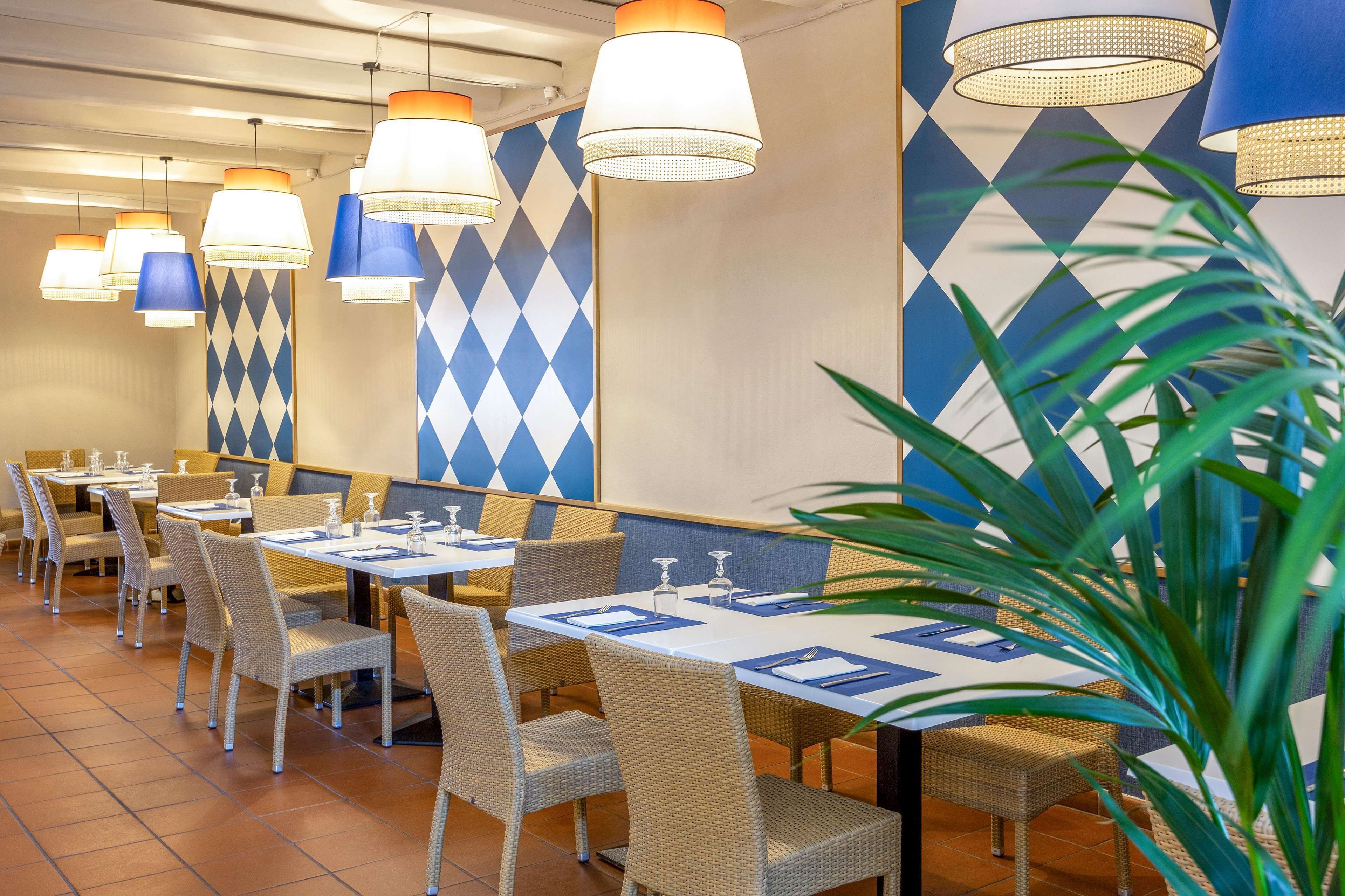 Um restaurante com cadeiras e mesas de vime, paredes com padrão azul e branco e luminárias pendentes.