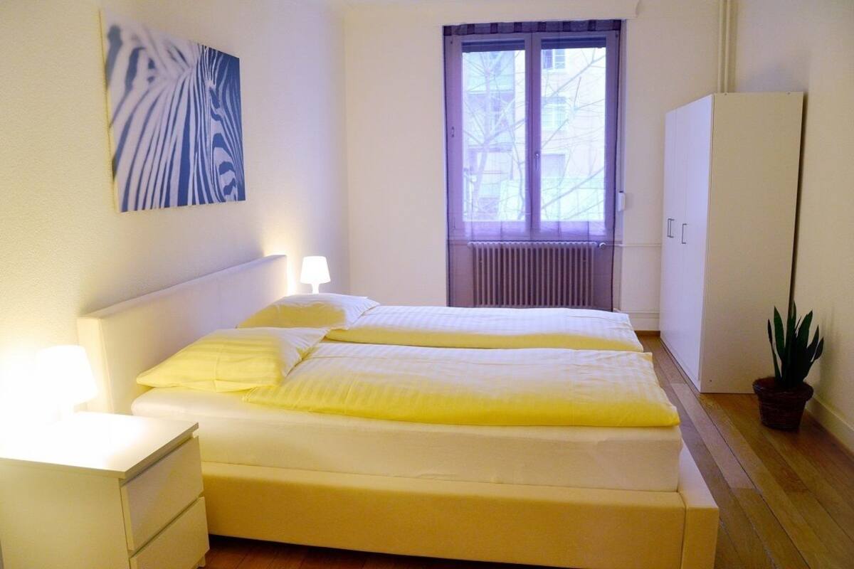 Quarto com cama, mesas de cabeceira, radiador, janela e planta em vaso.