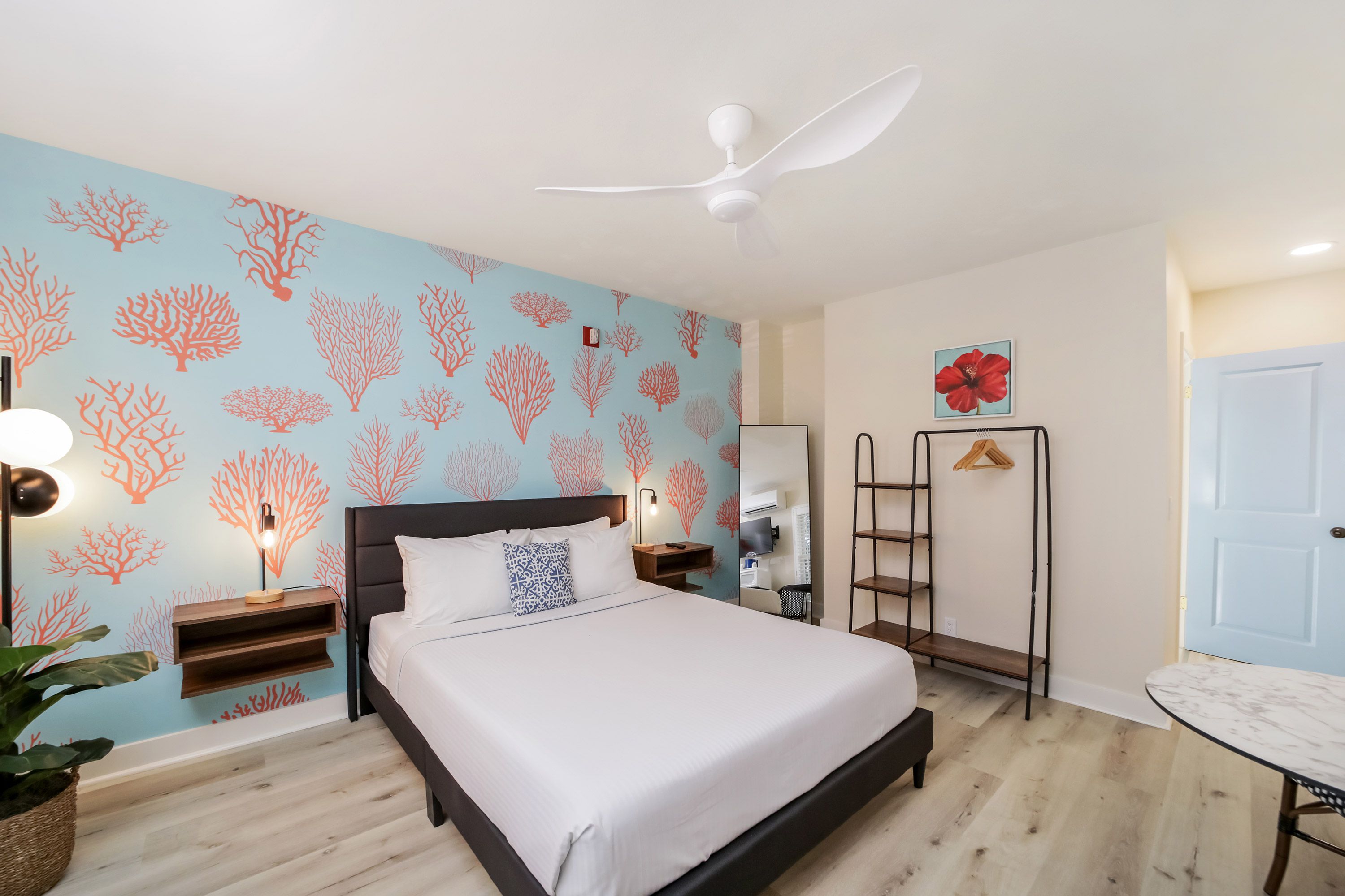 Quarto de hotel com cama, mesa de cabeceira, luminária, ventoinha de teto, espelho, televisão, guarda-roupa e uma pequena mesa.