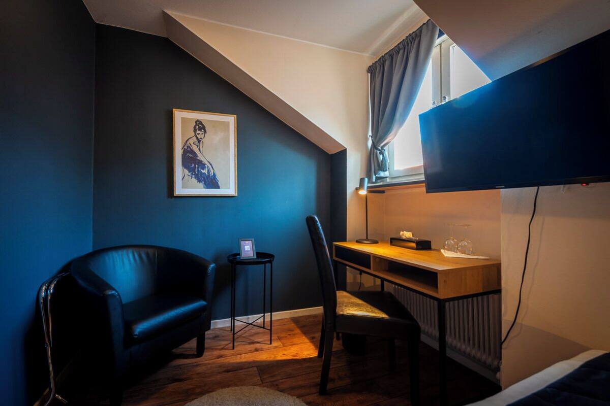 Um quarto pequeno com decoração escura, teto inclinado, uma poltrona de couro preto, uma escrivaninha de madeira, um quadro na parede e uma TV de tela plana fixada na parede.