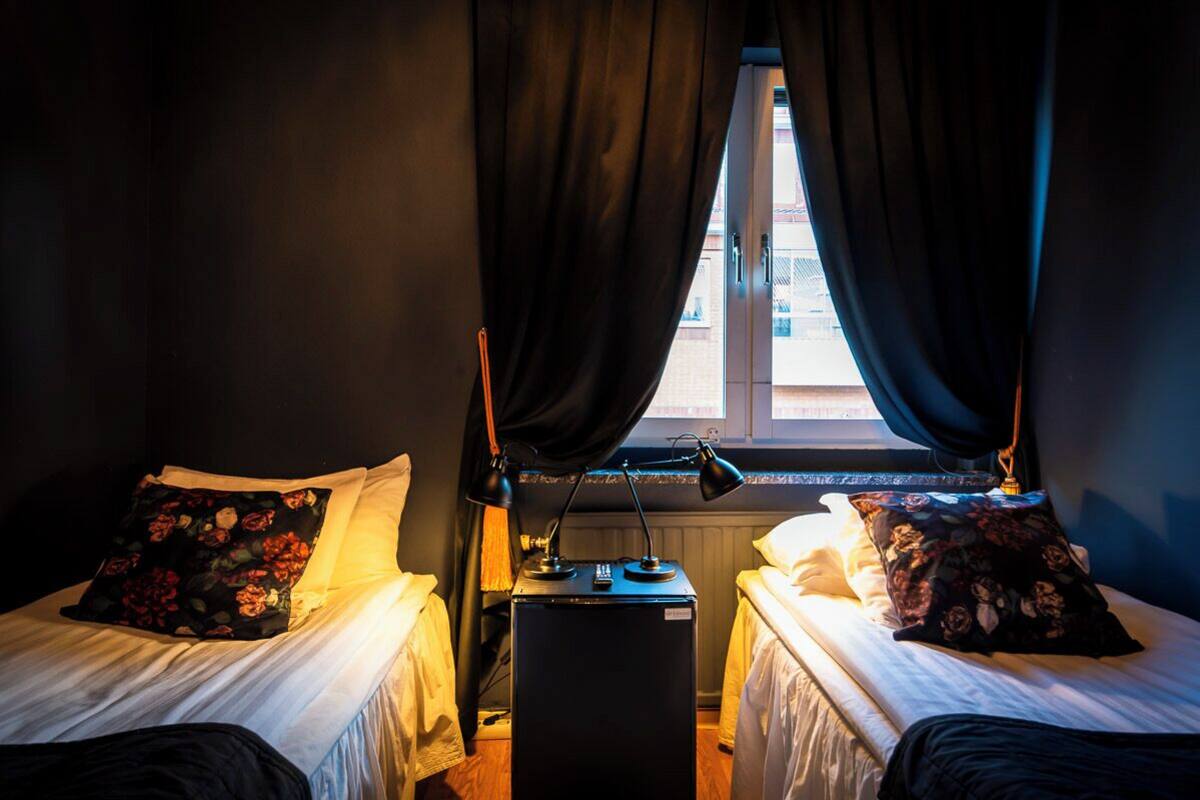Quarto pequeno com duas camas de solteiro, cada uma com almofadas florais, uma mesa de cabeceira com abajur e uma janela com cortinas.
