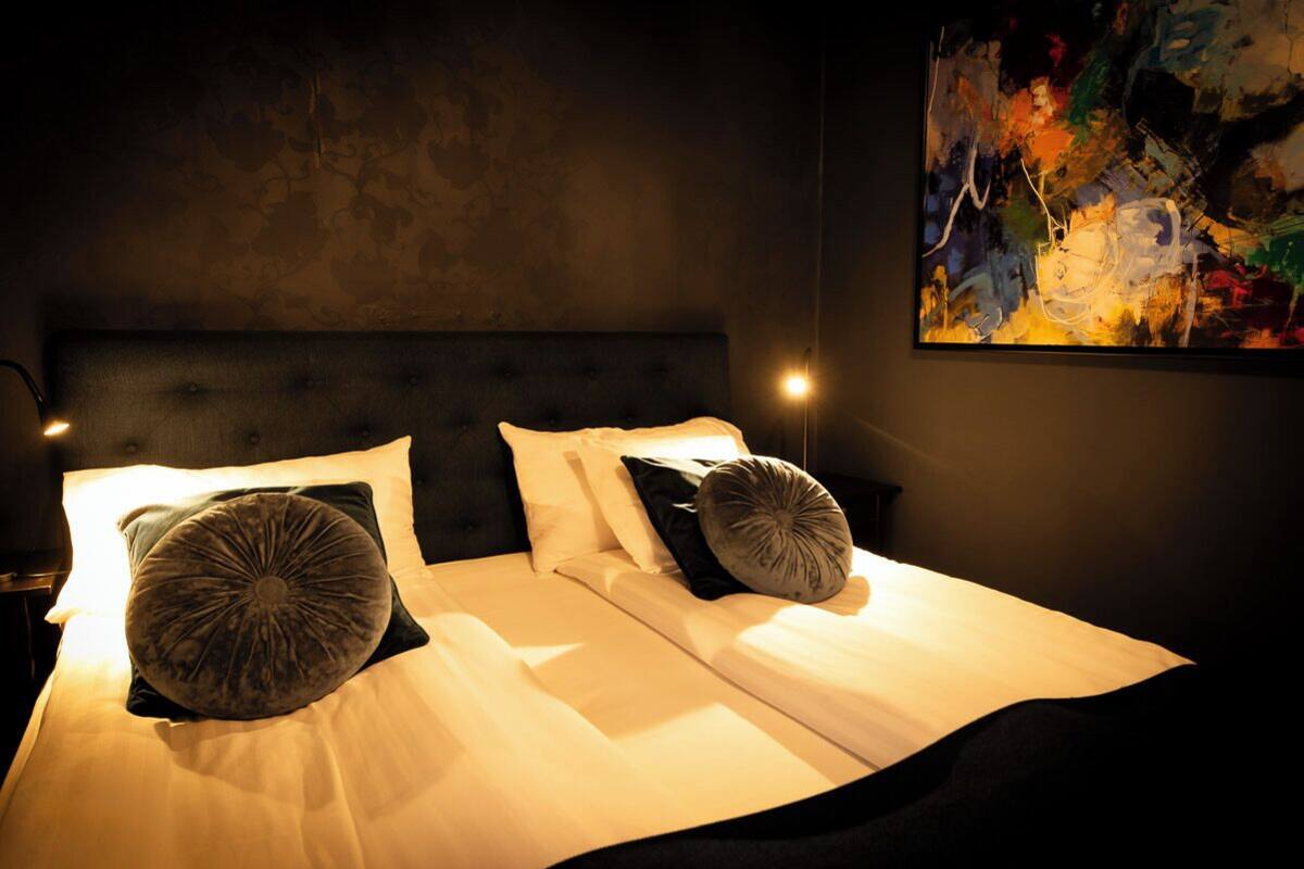 Uma cama bem arrumada com lençóis brancos e duas almofadas decorativas, uma cabeceira escura e uma pintura abstrata pendurada na parede.