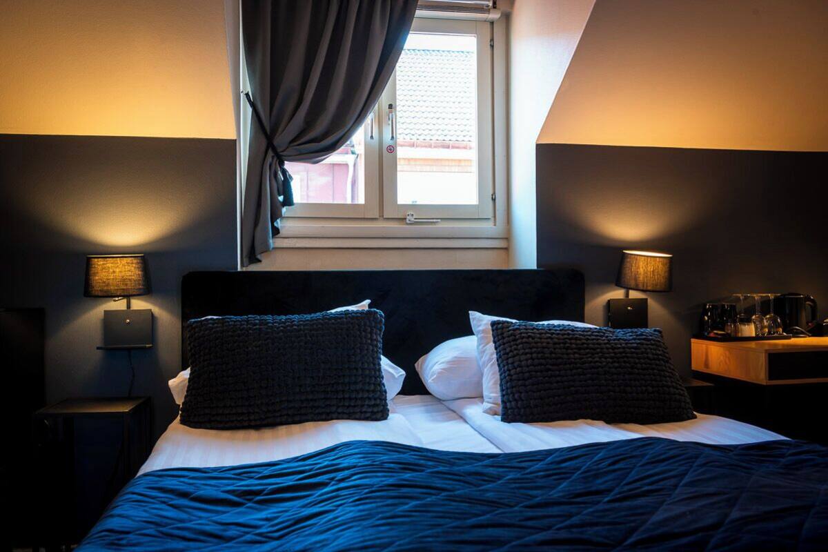 Quarto com cama, duas luminárias de cabeceira, janela com cortinas e mesa de cabeceira com cafeteira.
