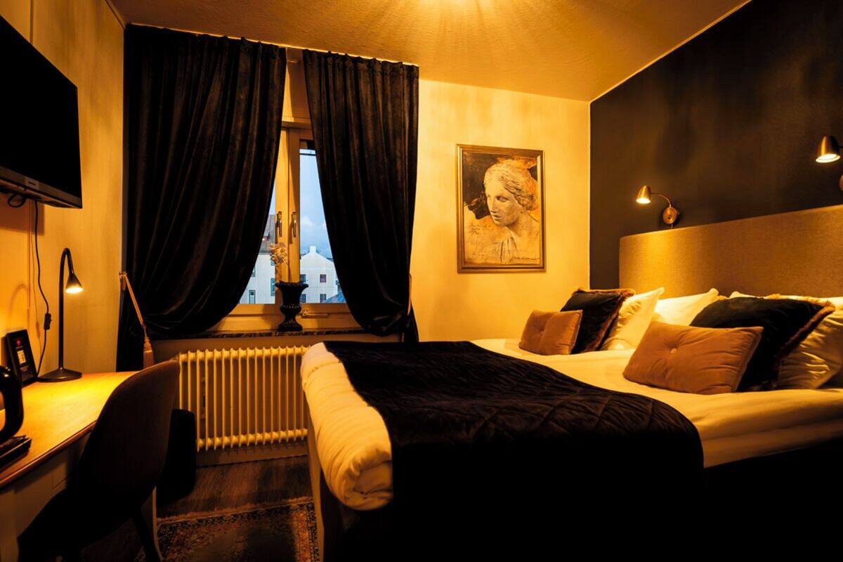 Quarto de hotel com cama, escrivaninha, cadeira, radiador, televisão e janela com cortinas.
