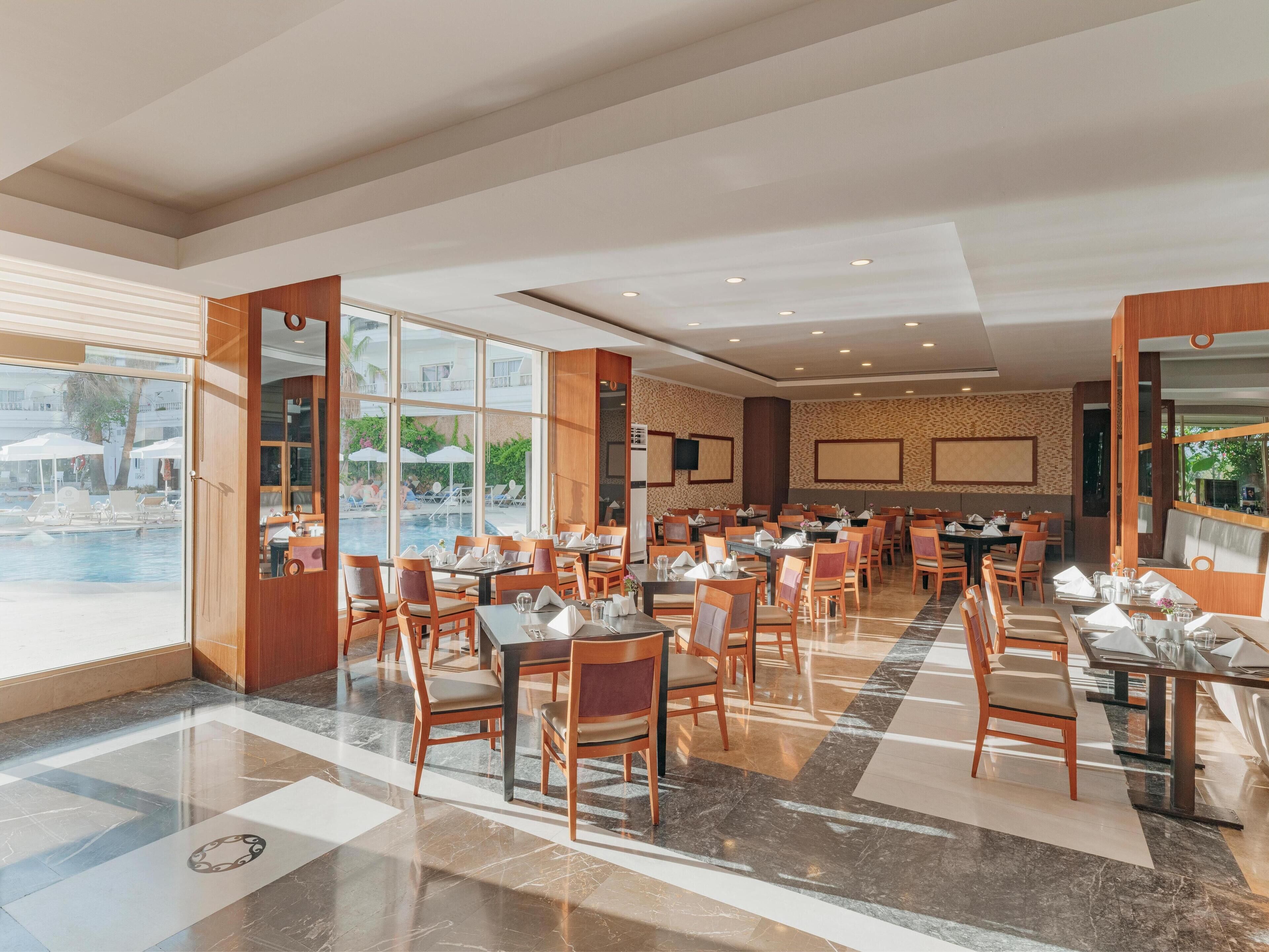 Um restaurante amplo com várias mesas e cadeiras, janelas grandes com vista para uma área de piscina e um design de interiores moderno.