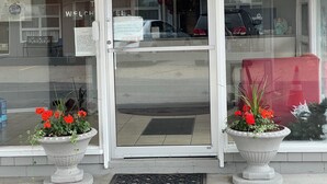 Entrada de hotel com porta de vidro, placa de 'Welcome' e dois vasos com plantas e flores vermelhas.