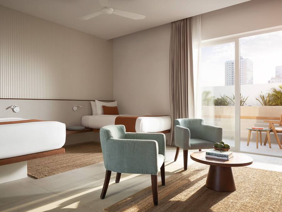 Quarto de hotel moderno com uma cama grande, duas poltronas, uma mesa de centro e varanda com vista para a cidade.