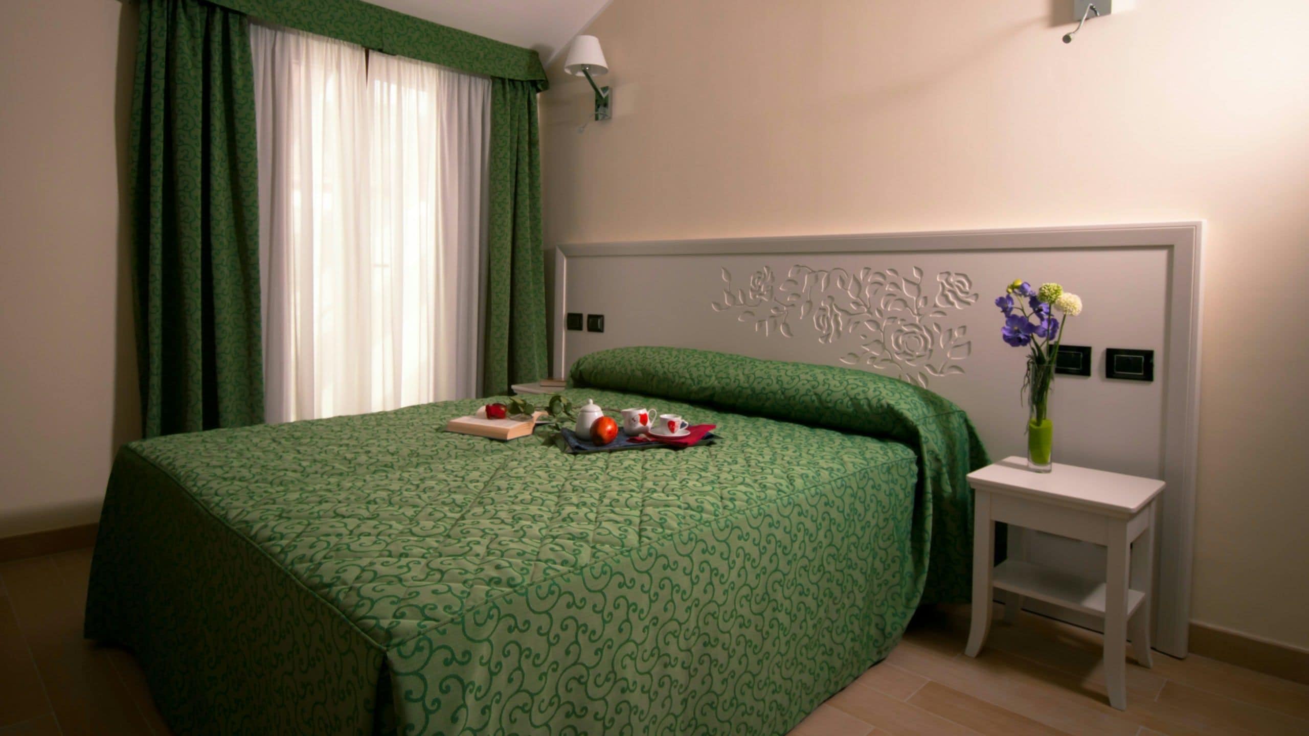 Quarto de hotel com colcha verde estampada, mesa de cabeceira com um vaso de flores e painel decorativo na parede.