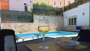 Um copo de vinho branco sobre uma mesa com um laptop e recipientes com petiscos, com vista para uma piscina, uma parede de pedra e uma cerca.