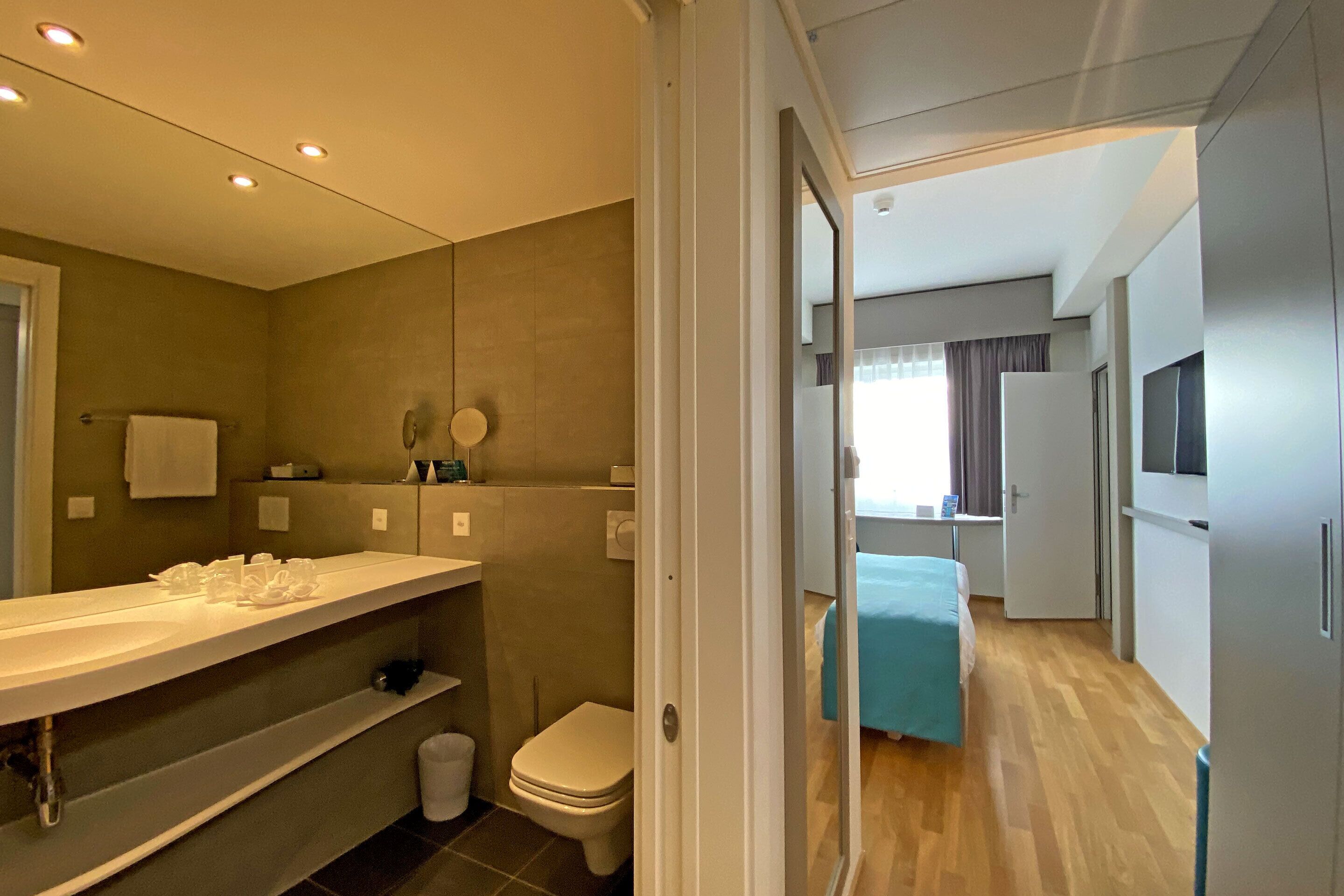 Uma casa de banho moderna com um espelho grande, sanita e lavatório. Existe uma área de duche com porta de vidro e um porta-toalhas. O quarto adjacente tem soalho em madeira e uma cama com colcha azul.