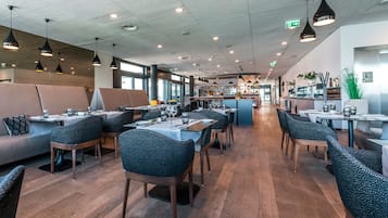 Interior de restaurante moderno com piso de madeira, cadeiras estofadas escuras e iluminação pendente.
