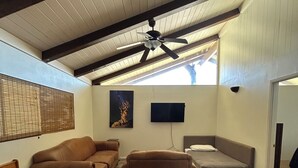 Um quarto amplo com teto alto, vigas de madeira, ventoinha no teto, uma janela grande, um sofá, um banco e uma TV fixada na parede.