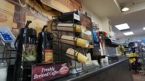 Balcão de café com exposição de máquinas de café e xícaras, um letreiro anunciando café fresco e uma grande placa com publicidade de café gourmet.