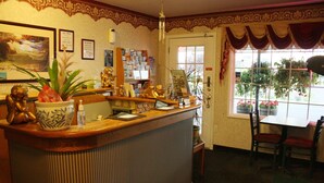 Uma área de recepção com balcão de madeira, quadros decorativos, uma janela com cortinas e uma mesa com cadeiras.