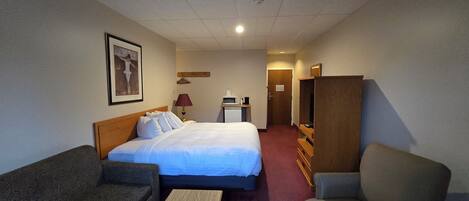 Quarto de hotel com cama, sofá, poltrona, escrivaninha, luminária, televisão e guarda-roupa.