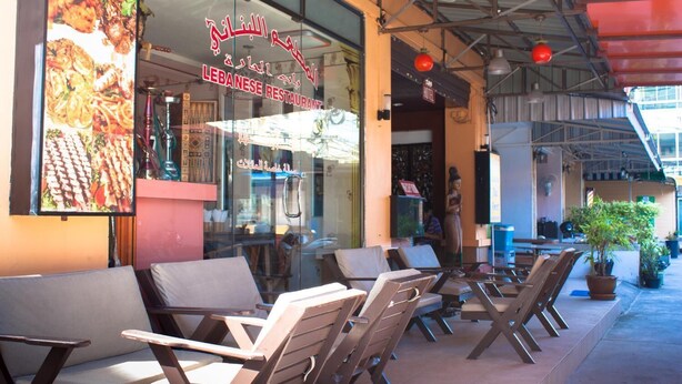 Uma área externa com mesas e cadeiras de madeira, uma parede de vidro com cardápio, e uma placa indicando restaurante libanês.