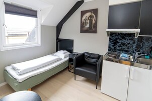 Quarto de hotel compacto com uma cama de solteiro, uma pequena cozinha, uma cadeira e um quadro na parede.