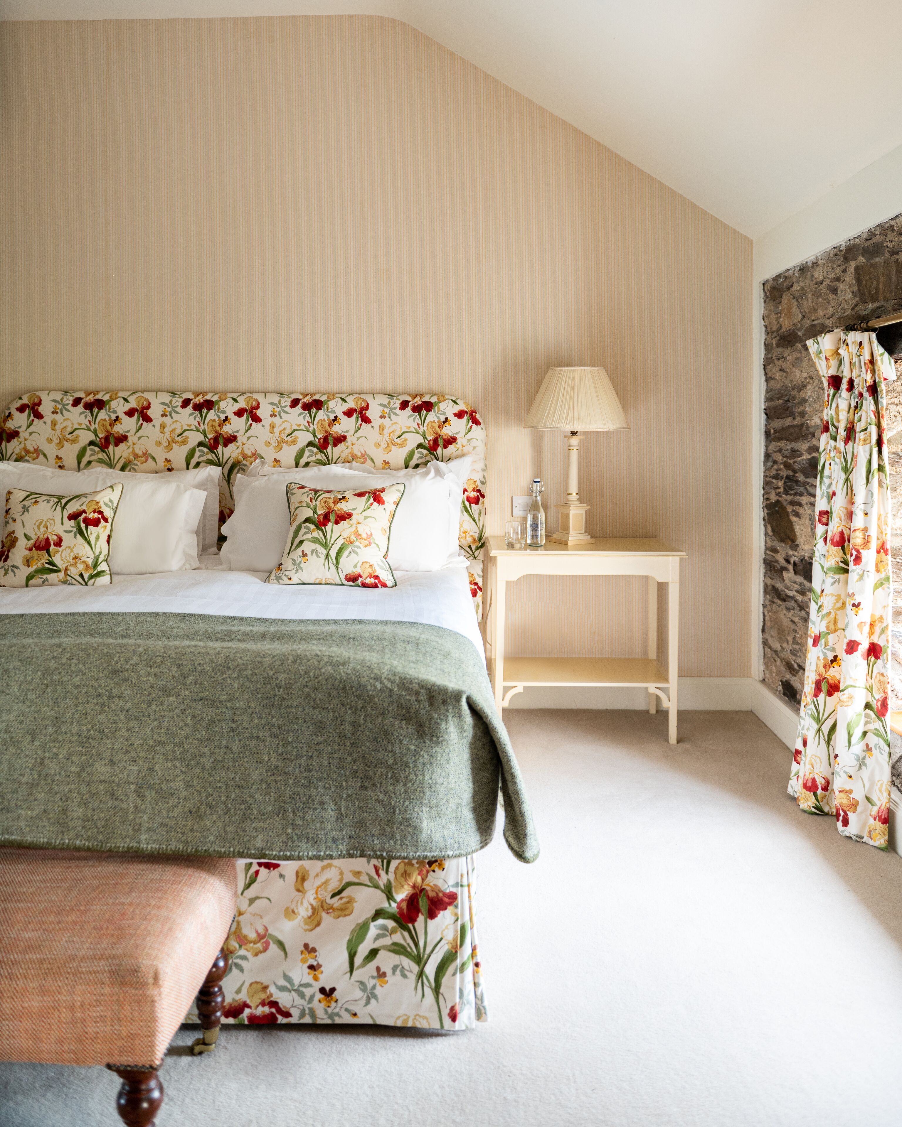 Quarto com colcha floral, mesa de cabeceira com abajur e parede de pedra.