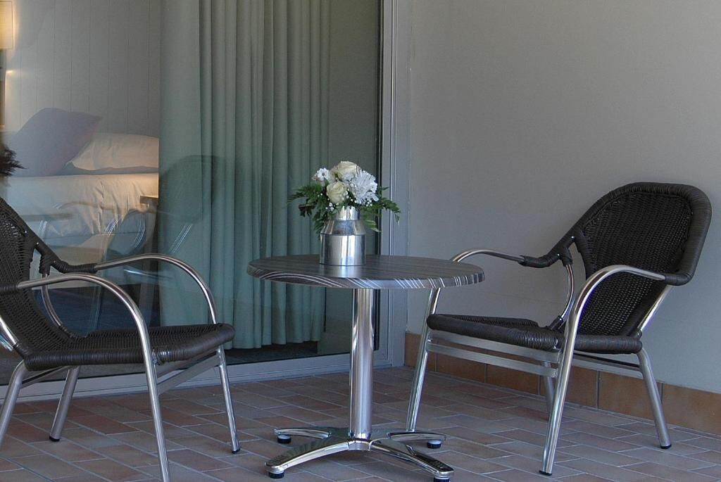 Uma varanda com duas cadeiras de vime, uma mesa redonda pequena com um vaso de flores e vista para um quarto com cama e cortinas.