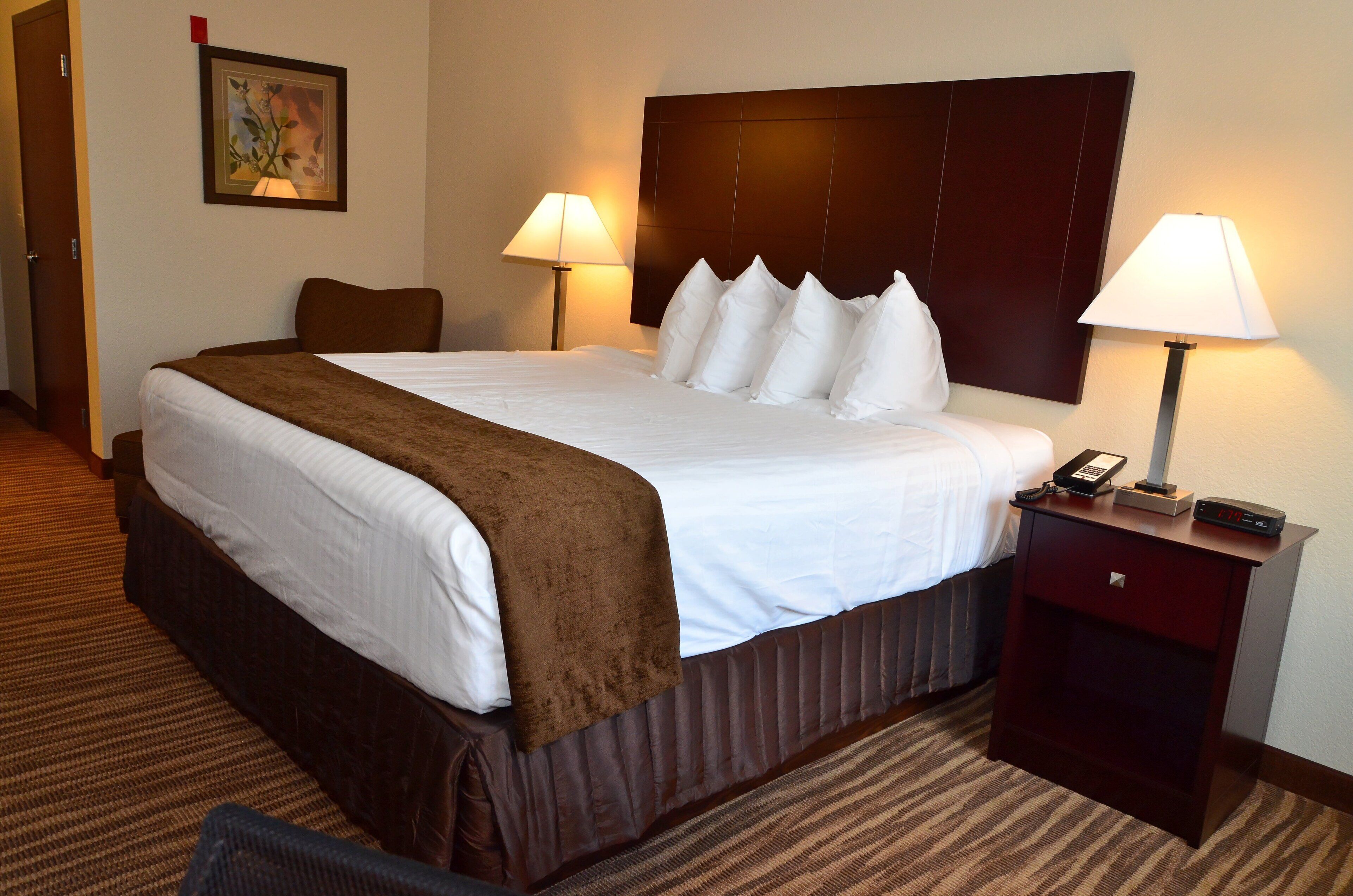 Quarto de hotel com uma cama grande, duas luminárias de cabeceira, mesa de cabeceira com telefone e relógio, e uma porta à esquerda.