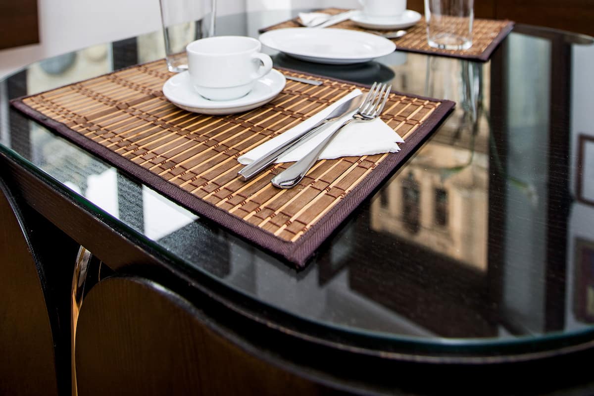Uma mesa de jantar com tampo de vidro, posta com pratos brancos, xícaras e talheres, e um copo de vidro transparente.
