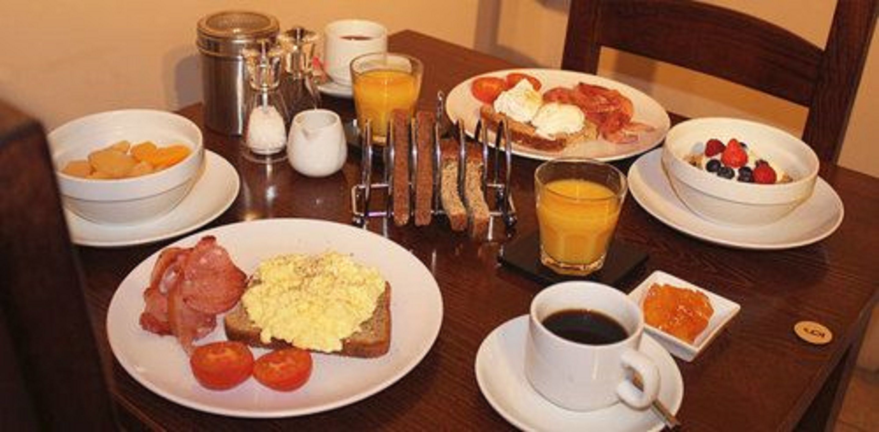 Uma mesa de café da manhã com pratos de ovos, bacon, torradas, tomates e uma tigela de frutas. Também há xícaras de café e suco.
