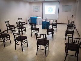 Uma sala de conferências com fileiras de cadeiras, uma tela de projetor e uma mesa coberta com toalha azul.