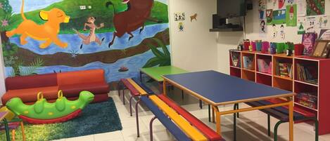 Uma sala de jogos infantil com um mural, móveis coloridos e uma estante de livros.