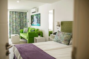 Quarto de hotel moderno com um sofá verde, uma cama com colcha roxa, um quadro de uma cascata e uma pequena mesa com uma planta.