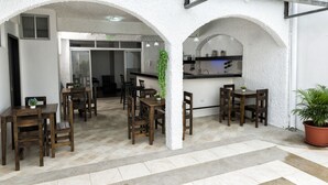 Um pátio externo com mesas e cadeiras de madeira, paredes brancas com textura e um vaso com planta.