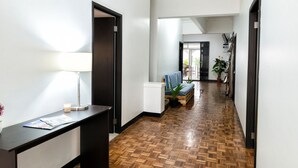 Corredor de hotel com chão em assoalho de madeira, um balcão de recepção com abajur, um banco e um vaso de planta.