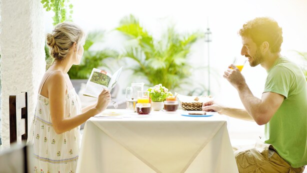 Uma mesa de jantar com toalha branca, uma cafeteira, um jarro de sumo de laranja e um pequeno vaso com flores.