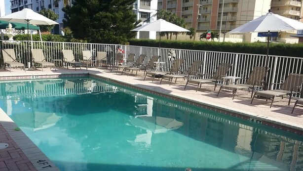 Uma piscina retangular com superfície de água azul translúcida, rodeada por uma cerca de metal branca. Há espreguiçadeiras e guarda-sóis ao longo da borda da piscina.
