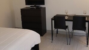 Quarto de hotel com uma cama, televisão sobre uma cômoda, escrivaninha com duas cadeiras e uma mesinha com duas taças de vinho.