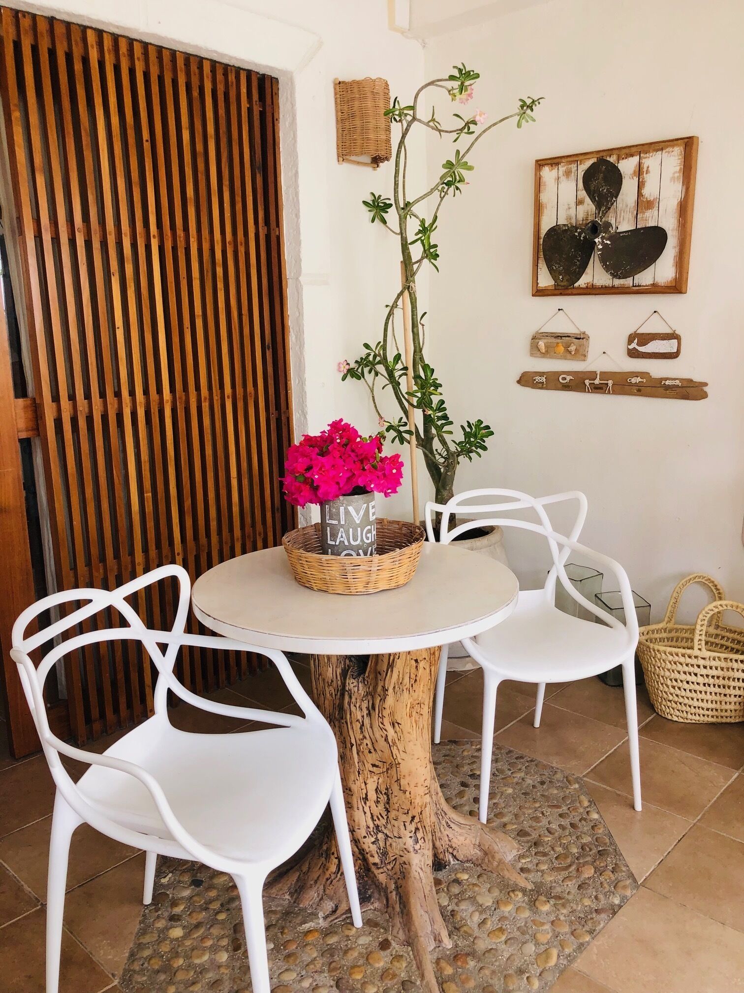 Uma mesa redonda com base de madeira, rodeada por cadeiras brancas, num quarto com uma porta de ripas de madeira e uma cesta trançada na parede.