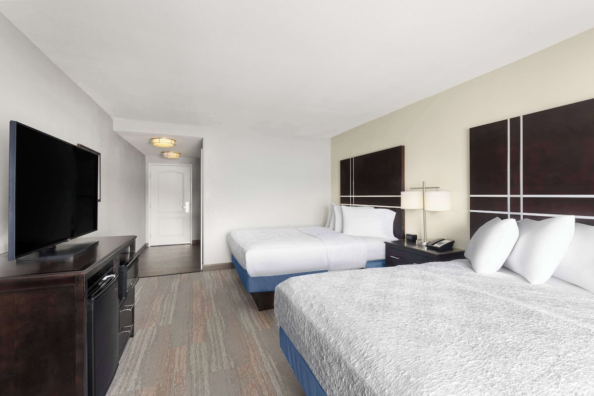 Quarto de hotel com uma cama grande, uma cama menor, uma TV, uma escrivaninha e um abajur.