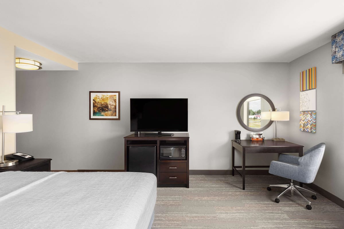 Quarto de hotel com cama, escrivaninha com computador, televisão, micro-ondas, cadeira e luminária.